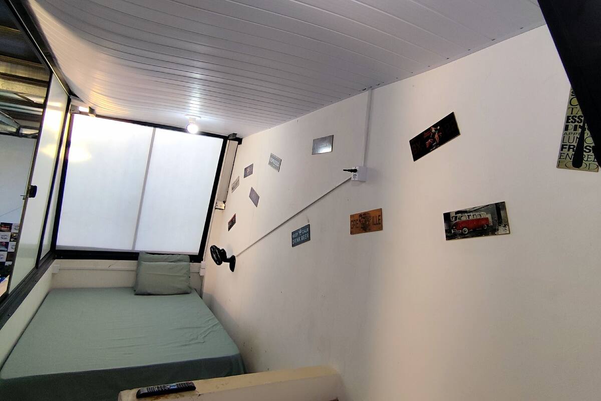 Quarto compacto com uma cama, um banco de madeira, uma luminária no teto e uma janela.
