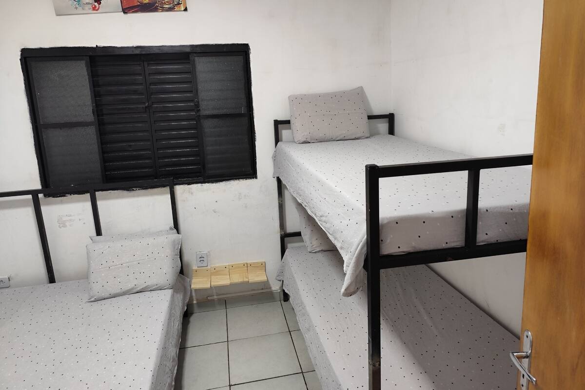 Um quarto tipo dormitório com beliches, uma janela com persianas, um interruptor de luz na parede e uma pequena prateleira com objetos.