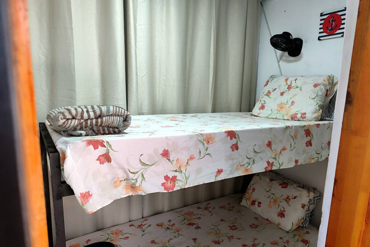 Uma cama beliche com roupa de cama florida, um cobertor dobrado e um ventilador fixado na parede.