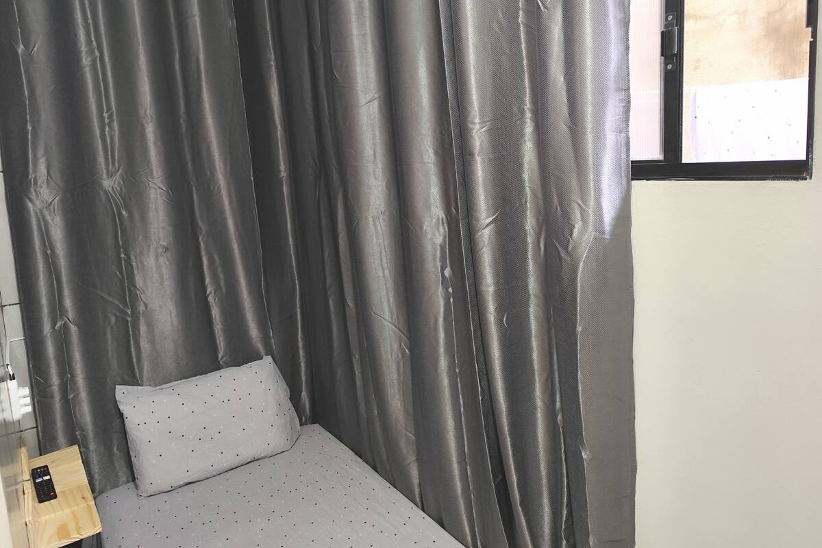 Quarto com cama, cortinas cinzas, uma janela e um controle remoto sobre uma superfície de madeira.