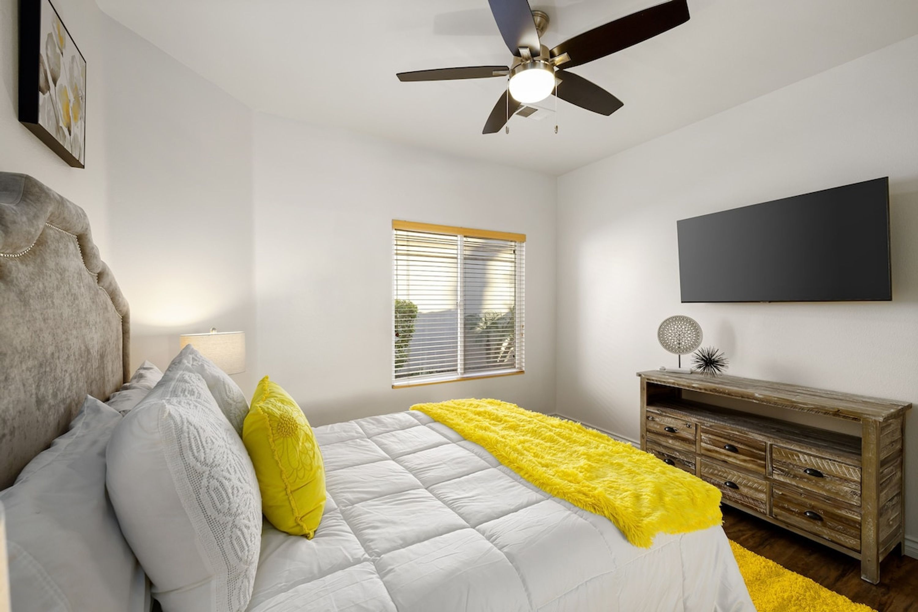 Quarto com cama, ventilador de teto, janela com persianas, televisão na parede e cômoda de madeira.