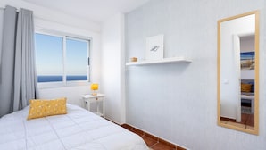 Quarto com cama, mesa de cabeceira, janela com cortinas, prateleira na parede com um quadro e espelho.