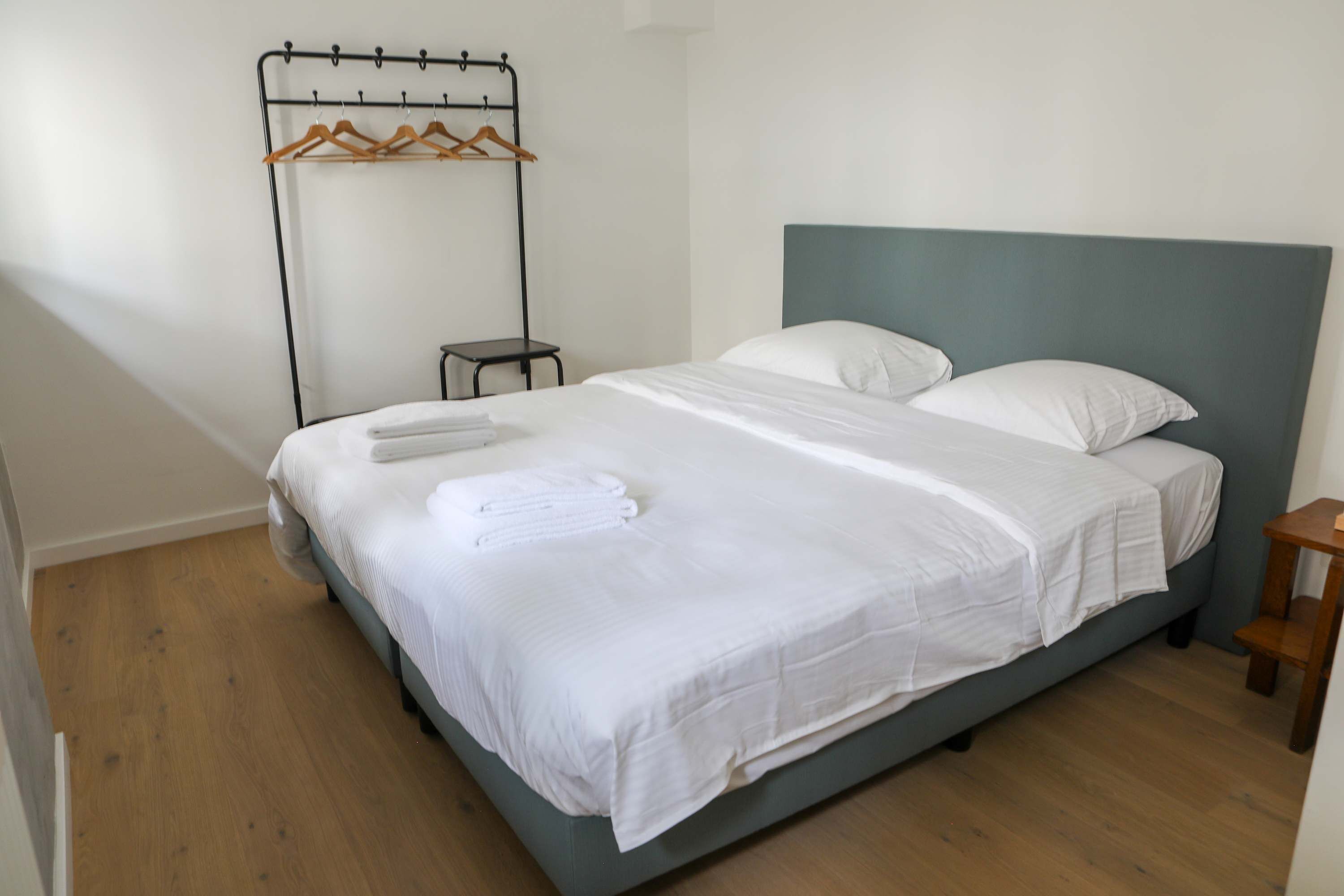 Uma cama bem arrumada com lençóis brancos e cabeceira cinza, um cabideiro de madeira com roupas e um pequeno banquinho ao lado da cama.