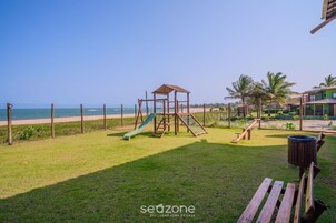 Um parquinho com escorrega e estruturas de escalada, cercado por uma grade, com uma praia e o oceano ao fundo.