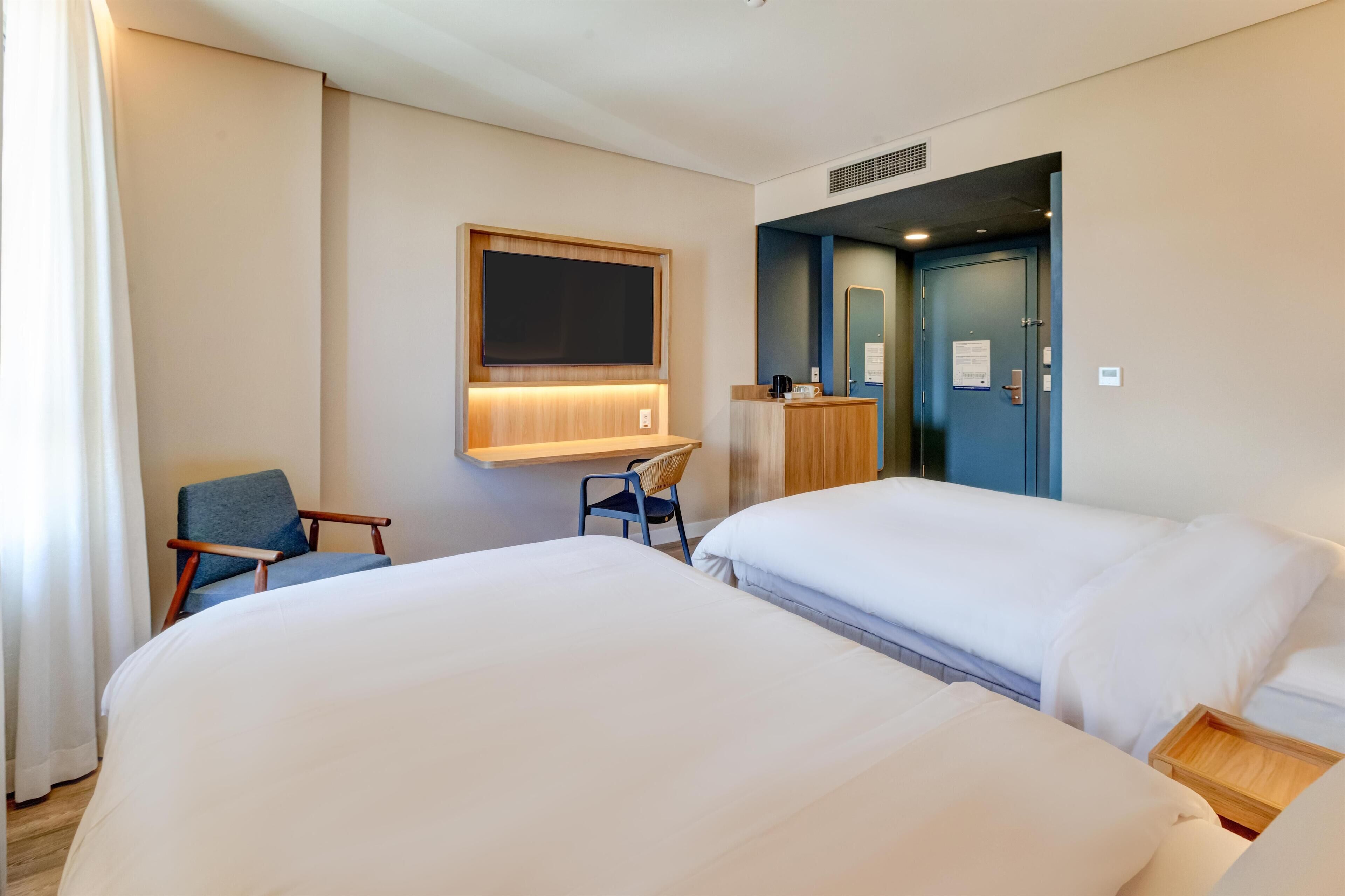 Quarto de hotel com duas camas, uma escrivaninha, uma cadeira, uma TV fixada na parede e uma porta para o banheiro.