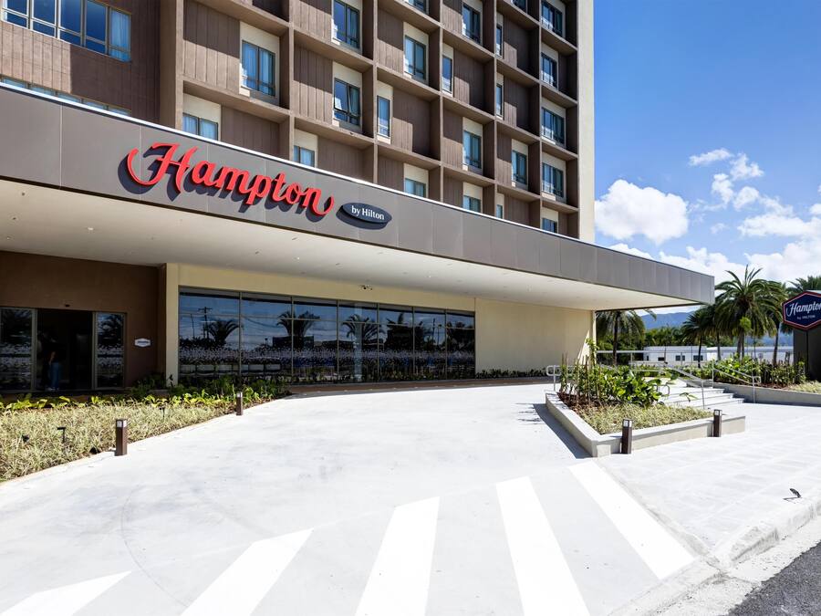 Entrada de um hotel Hampton by Hilton com design moderno, grandes janelas e céu claro.