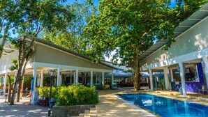 Uma área junto à piscina com espaço de estar coberto, piscina e vegetação exuberante.