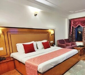 Quarto de hotel com uma cama grande, cabeceira de madeira, mesa de cabeceira, uma cadeira e janela com cortinas.