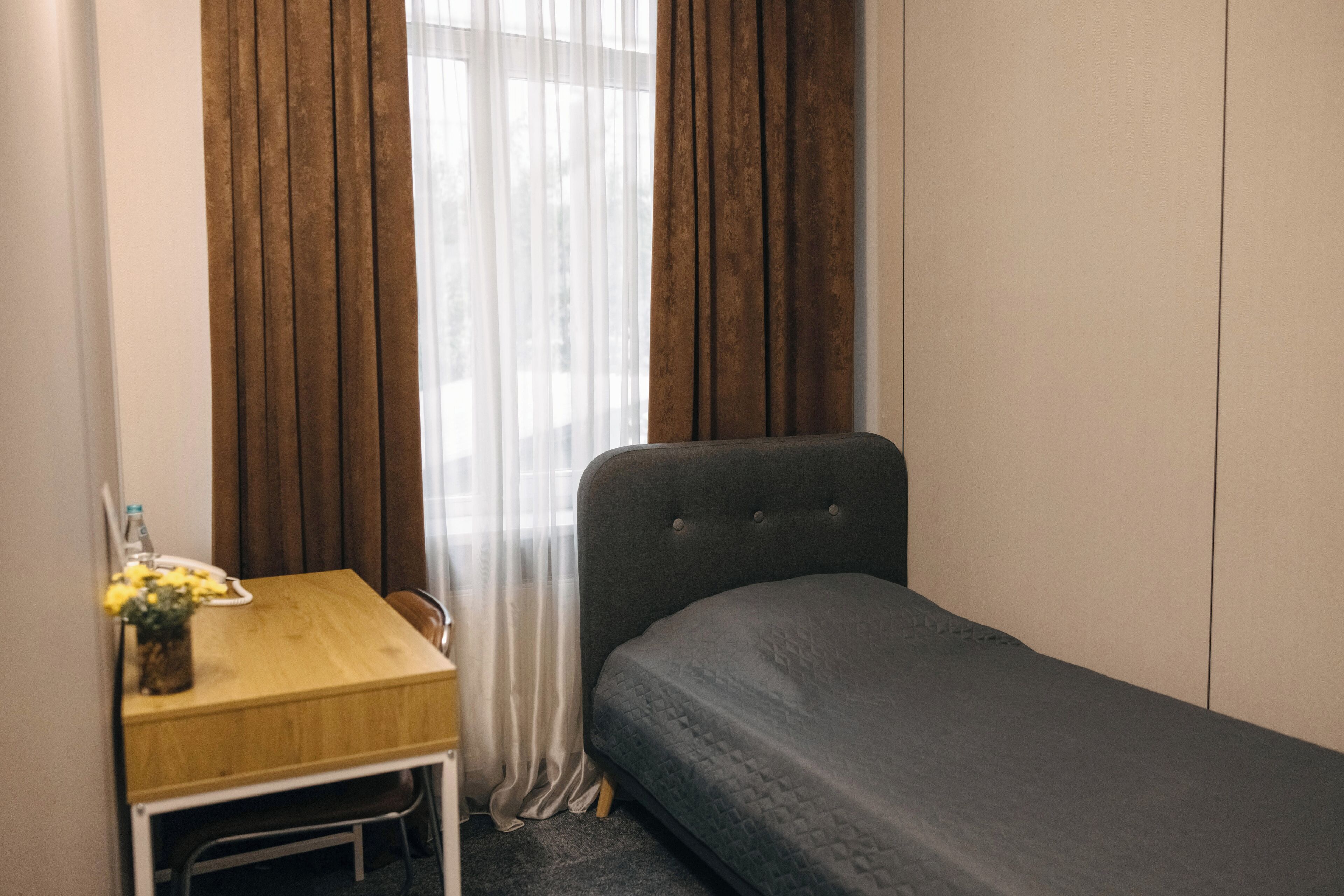 Quarto de hotel pequeno e bem arrumado, com uma cama de solteiro, uma escrivaninha de madeira com cadeira e uma janela com cortinas.