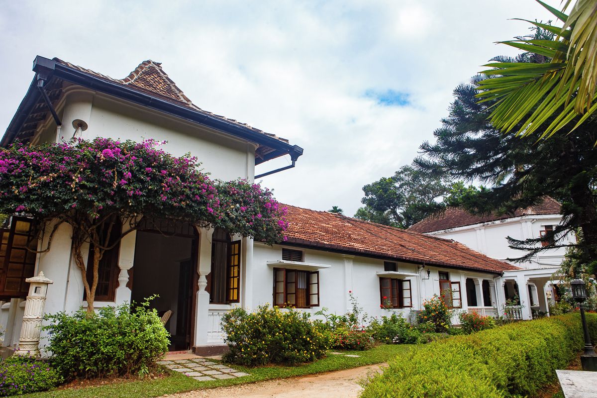 Um edifício branco em estilo colonial, com telhado de cerâmica, rodeado por vegetação exuberante e plantas floridas.