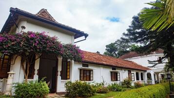 Um edifício branco em estilo colonial, com telhado de cerâmica, rodeado por vegetação exuberante e plantas floridas.