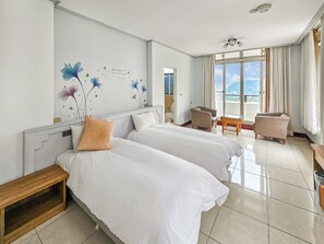 Quarto de hotel com duas camas, um banquinho de madeira, uma mesinha, uma cadeira e vista para o exterior.