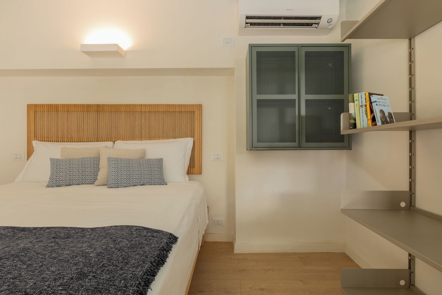 Quarto com cabeceira de madeira, cama com roupa de cama branca e almofadas estampadas, manta cinza, ar condicionado fixado na parede e um armário verde com livros.