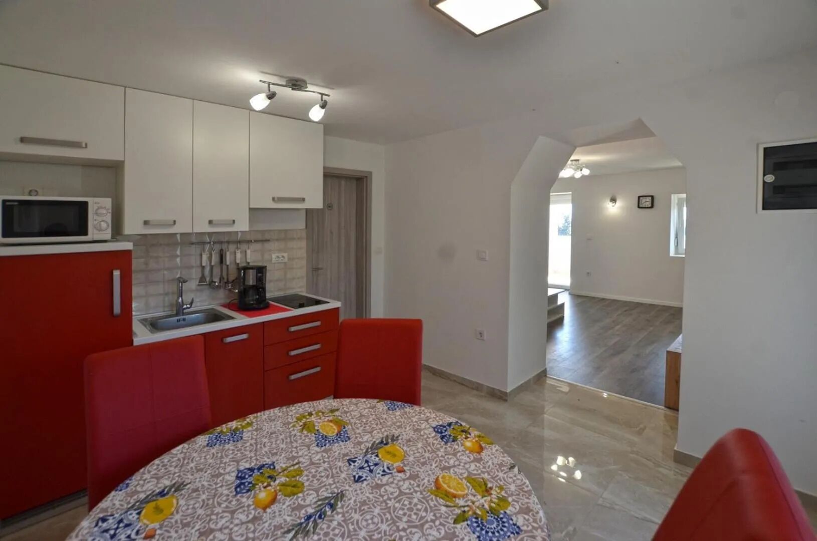 Cozinha compacta com armários brancos, bancada vermelha e toalha de mesa estampada.