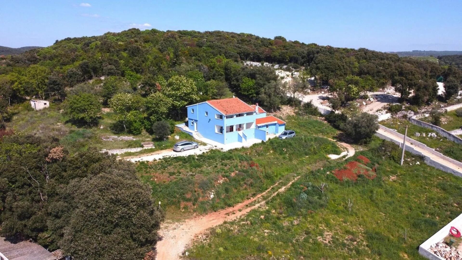 Uma casa azul de dois andares com telhado vermelho, rodeada por vegetação e um caminho de terra.