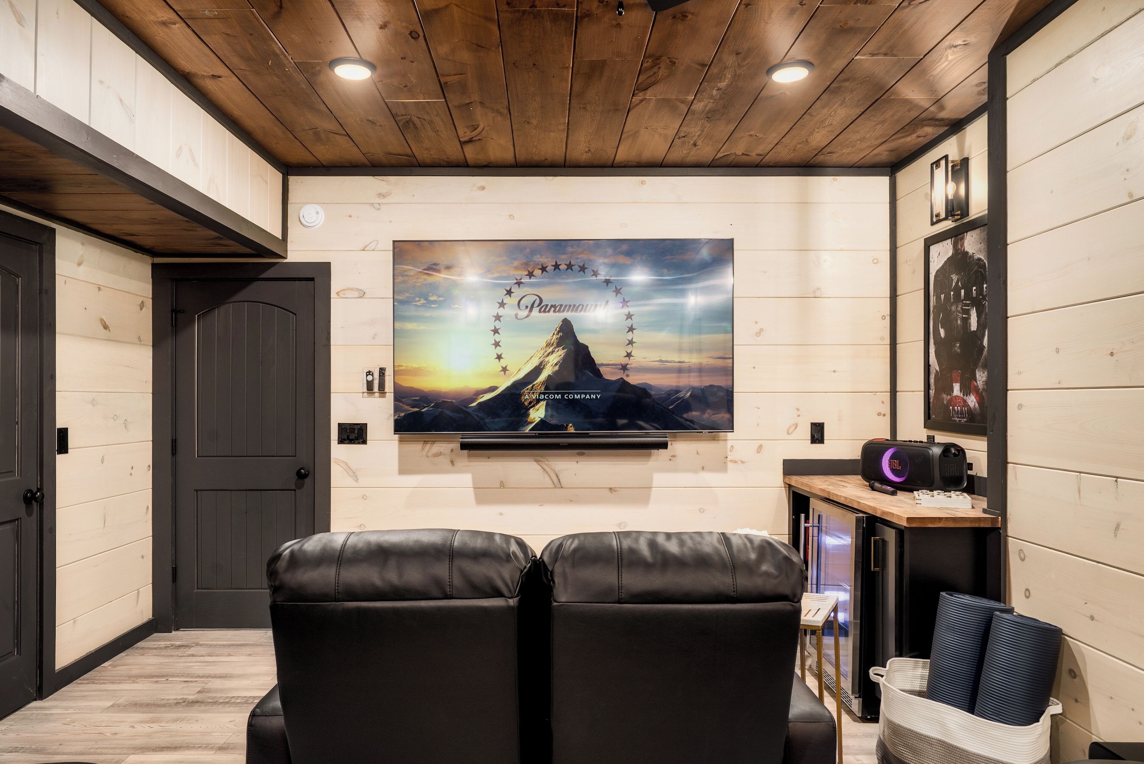Quarto com televisão na parede exibindo um pôster de filme Paramount, teto de madeira e área de estar com dois sofás de couro preto.
