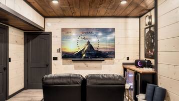 Quarto com televisão na parede exibindo um pôster de filme Paramount, teto de madeira e área de estar com dois sofás de couro preto.