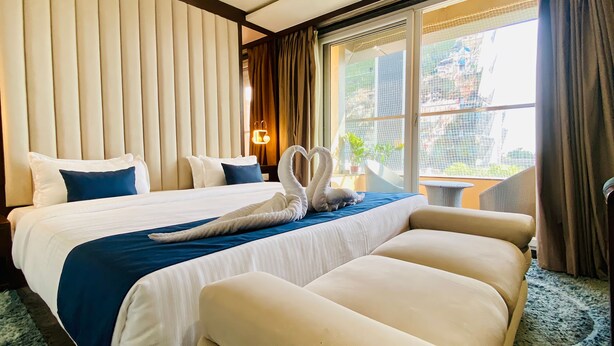 Quarto de hotel com uma cama grande, duas almofadas em forma de cisne, vista da cidade pela janela e uma varanda com uma cadeira.