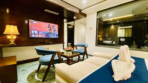 Quarto de hotel moderno com televisão de tela plana, duas poltronas azuis, um sofá bege, uma mesinha de centro com um vaso de flores e uma janela grande com vista para o interior.