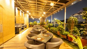 Restaurante no terraço com mesas ao ar livre, plantas em vasos e vista para a cidade à noite.