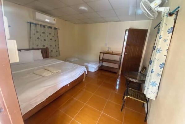 Um quarto com uma cama, uma cadeira, uma mesinha e um ventilador.