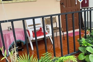 Uma varanda com guarda-corrimão preto, uma cadeira branca com almofada vermelha, uma mesa redonda pequena com toalha rosa e plantas em vasos.
