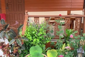 Varanda de madeira com grade decorativa, rodeada por diversas plantas e flores.