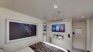 Quarto de hotel com uma cama grande, uma TV fixada na parede, um painel de informações digital e uma porta com câmera de segurança.