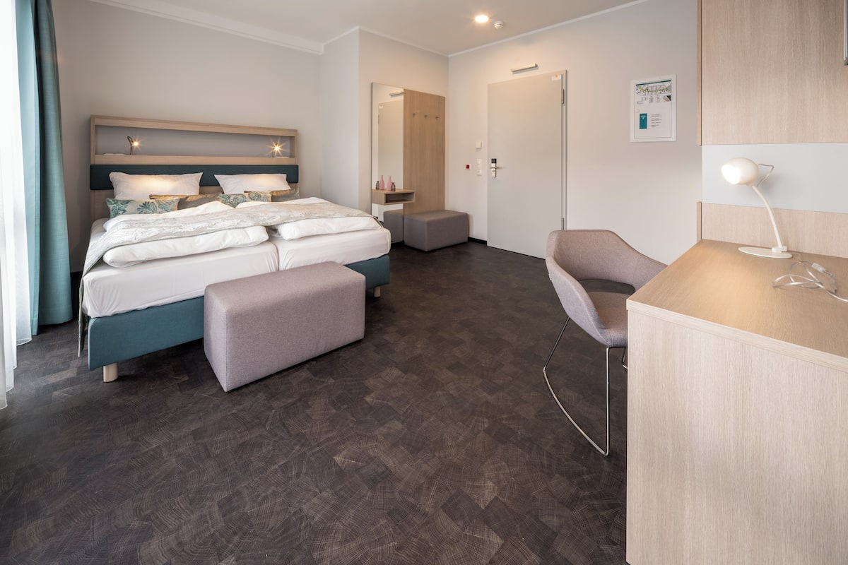 Quarto de hotel moderno com uma cama grande, uma escrivaninha e uma cadeira. Há uma porta e uma pequena mesa com uma luminária.