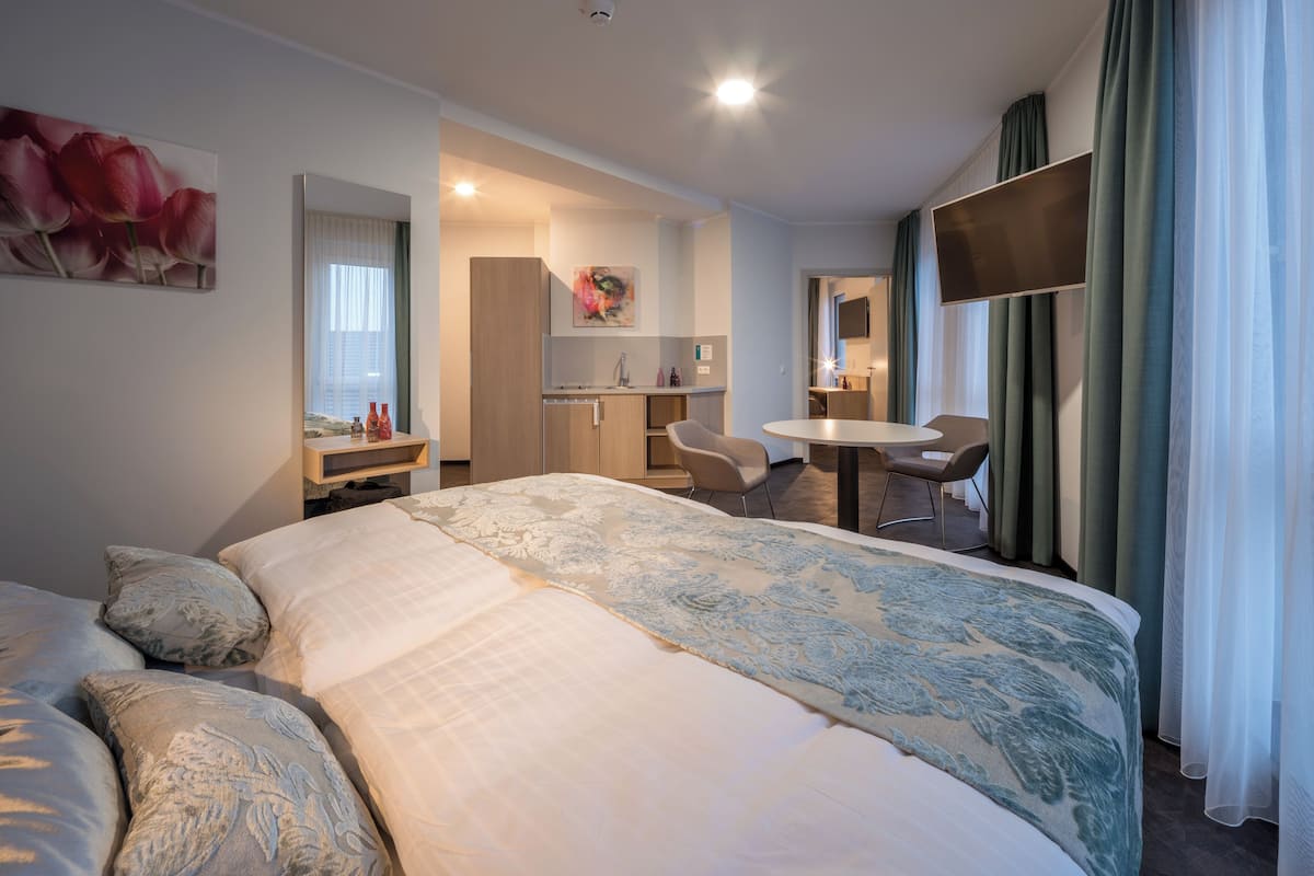 Quarto de hotel com uma cama grande, uma escrivaninha com cadeira, uma kitchenette pequena e um banheiro.