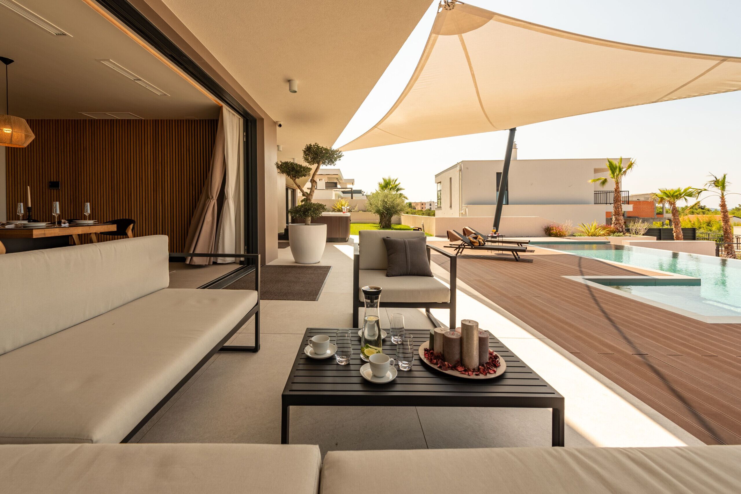 Uma área externa com mobiliário, incluindo piscina, sofá, mesa com bebidas e um grande guarda-sol.