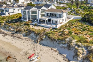 Uma casa moderna de dois andares com uma varanda grande, localizada em uma praia arenosa com uma escadaria que dá acesso à praia.