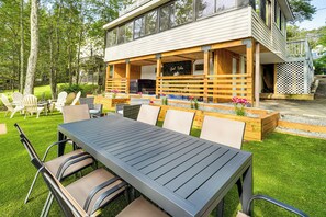 Uma área de refeições ao ar livre com uma mesa comprida e cadeiras, um deck de madeira e vista para uma casa moderna com deck e lareira.