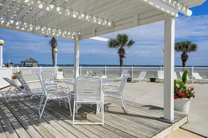 Área de estar coberta ao ar livre, com cadeiras de metal branco e uma mesa, com vista para uma praia com palmeiras e um cercado branco.