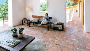 Um quarto espaçoso com teto de madeira, chão de ladrilho de pedra, um banco, uma mesa com livros e plantas, e vista para o verde lá fora.