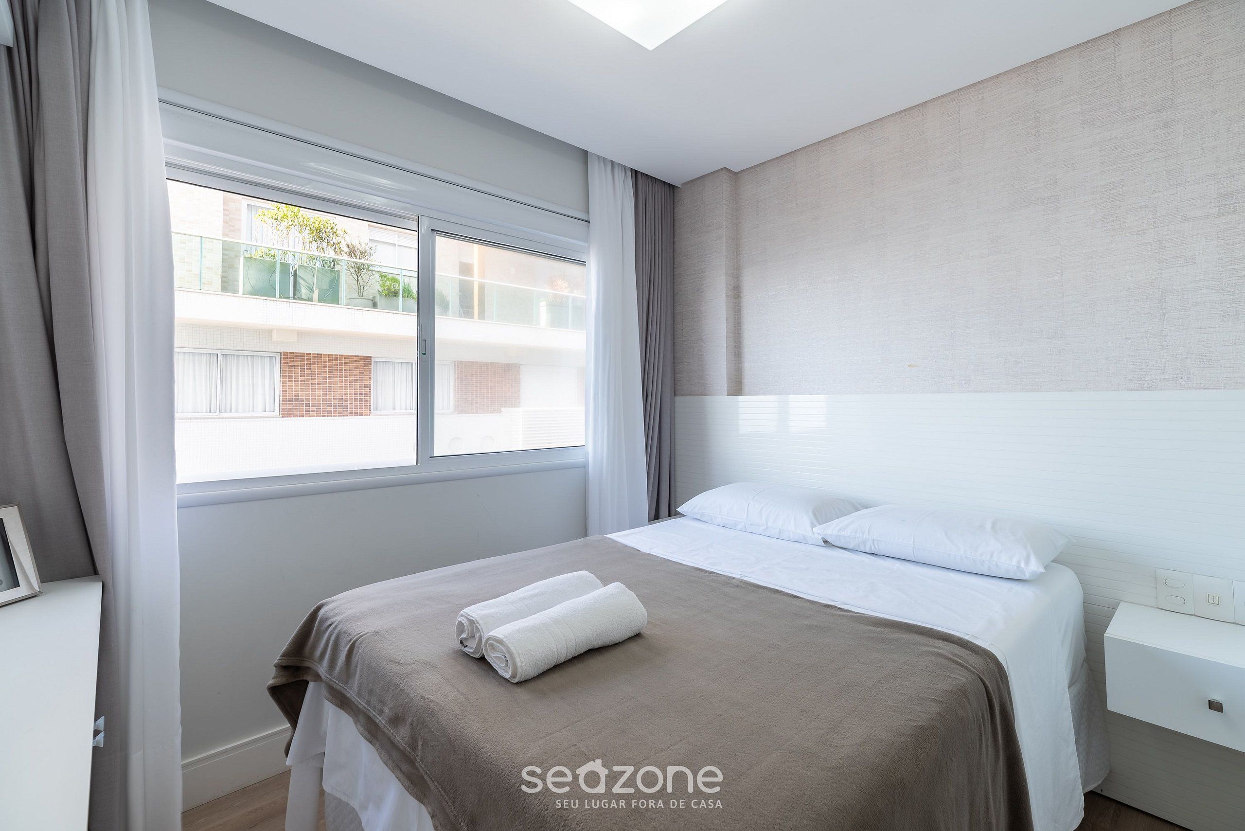 Quarto de hotel moderno com uma cama grande, uma mesa de cabeceira e uma janela com cortinas.