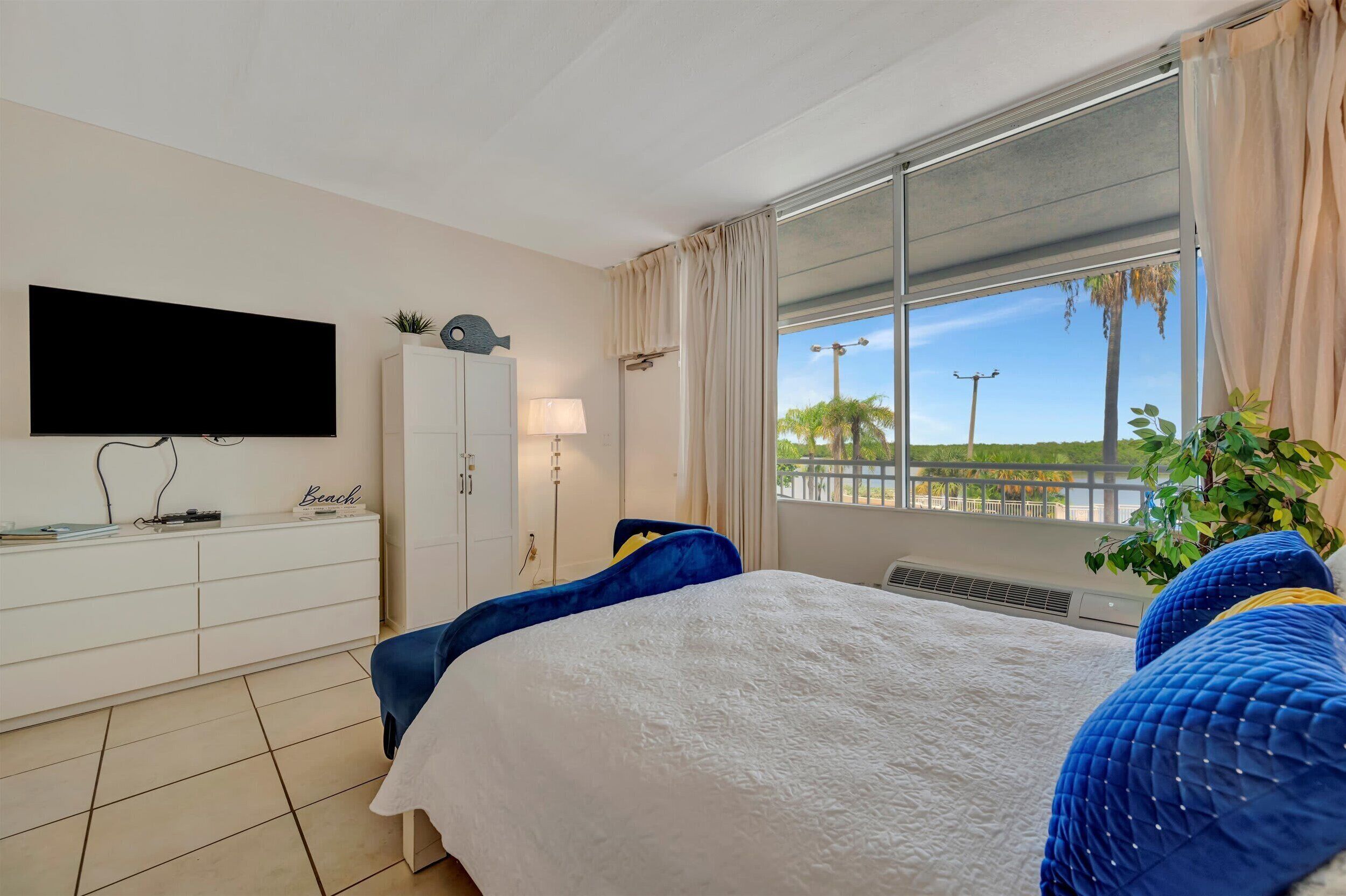 Quarto de hotel com uma cama, televisão, escrivaninha e janela com vista para palmeiras.