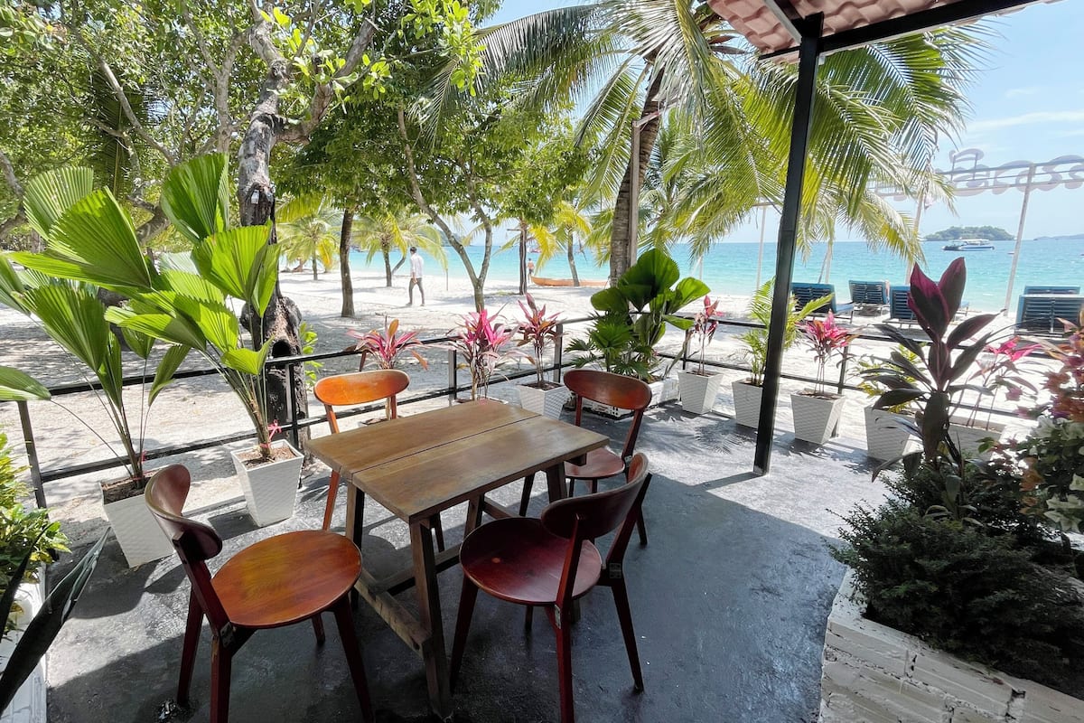 Conjunto de mesa e cadeiras de madeira ao ar livre, com plantas em vasos, vista para a praia e uma palmeira.