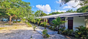 Fileira de edifícios térreos com chão arenoso, cercada por vegetação tropical e árvores.