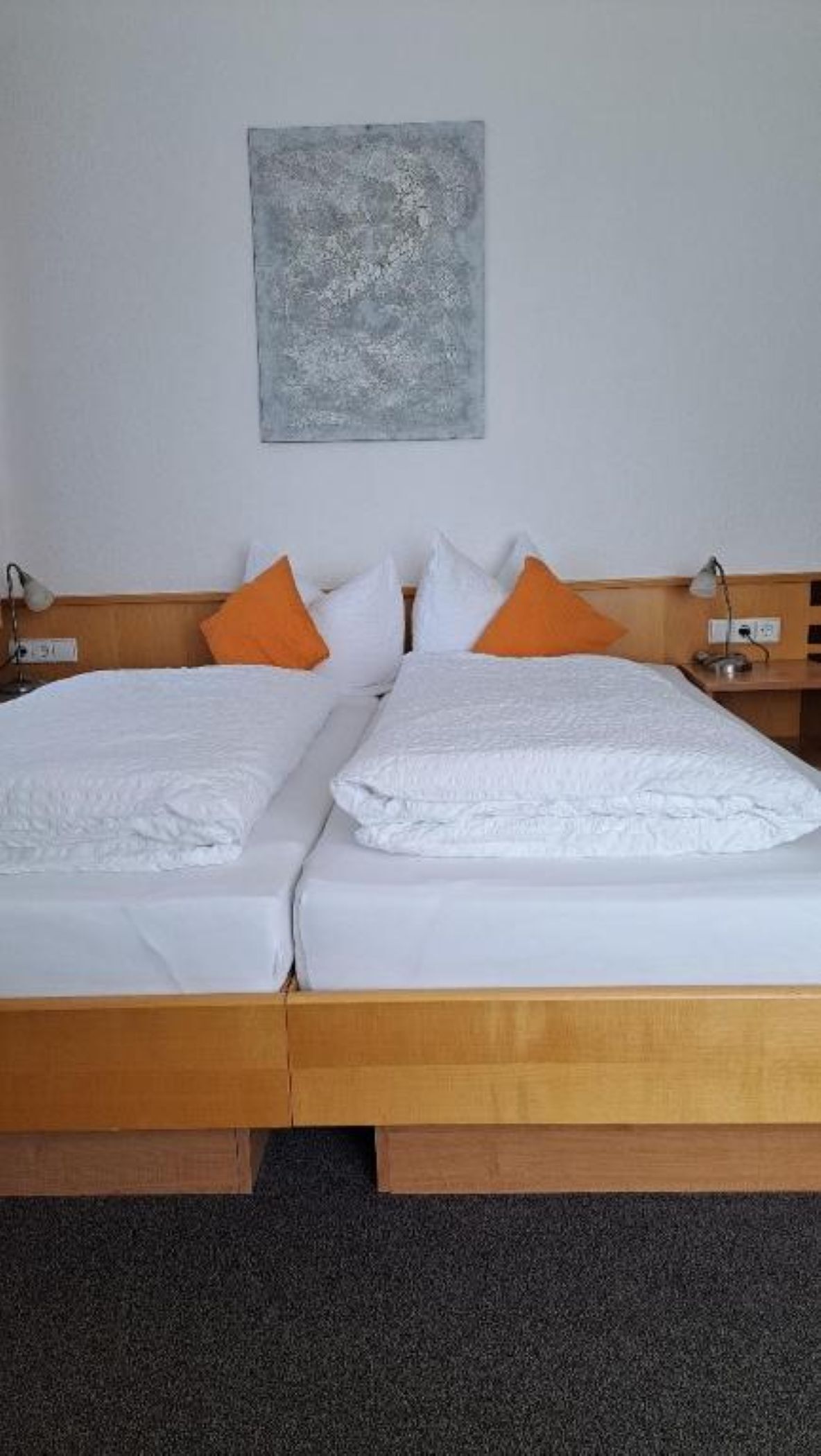 Uma cama de casal com lençóis brancos e almofadas laranja, uma cabeceira de madeira, uma pequena mesa de cabeceira com um abajur e um quadro abstrato cinza na parede.