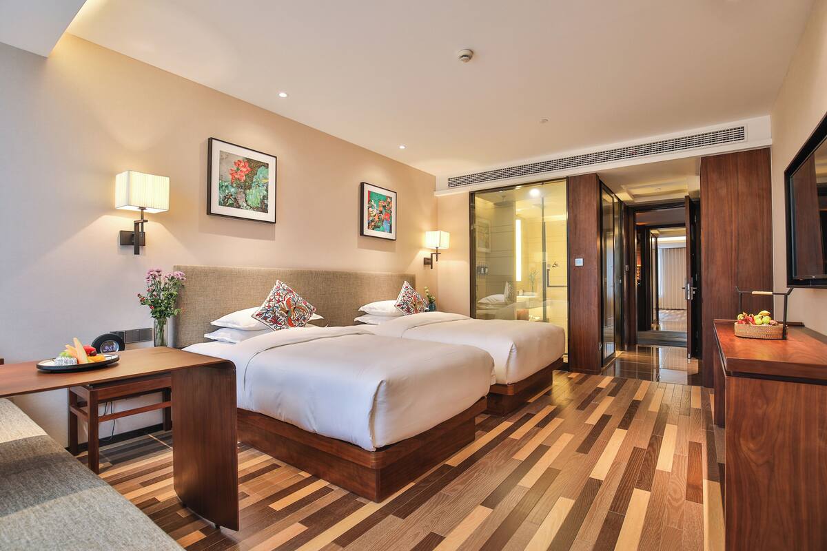 Quarto de hotel com duas camas, uma escrivaninha e um banheiro visível através de uma porta aberta.