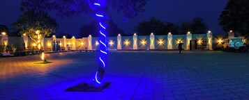 Área externa noturna com instalação de iluminação decorativa, calçada pavimentada e um edifício com detalhes iluminados na fachada.