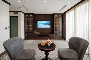 Um hall de hotel com uma televisão de tela plana grande, dois poltronas, um piso estampado e uma pequena mesa com um vaso e frutas.