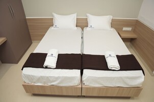 Quarto de hotel com duas camas de solteiro, cada uma com colchão branco e cabeceira marrom. Há um guarda-roupa de madeira à esquerda e uma mesa de cabeceira com uma luminária à direita.