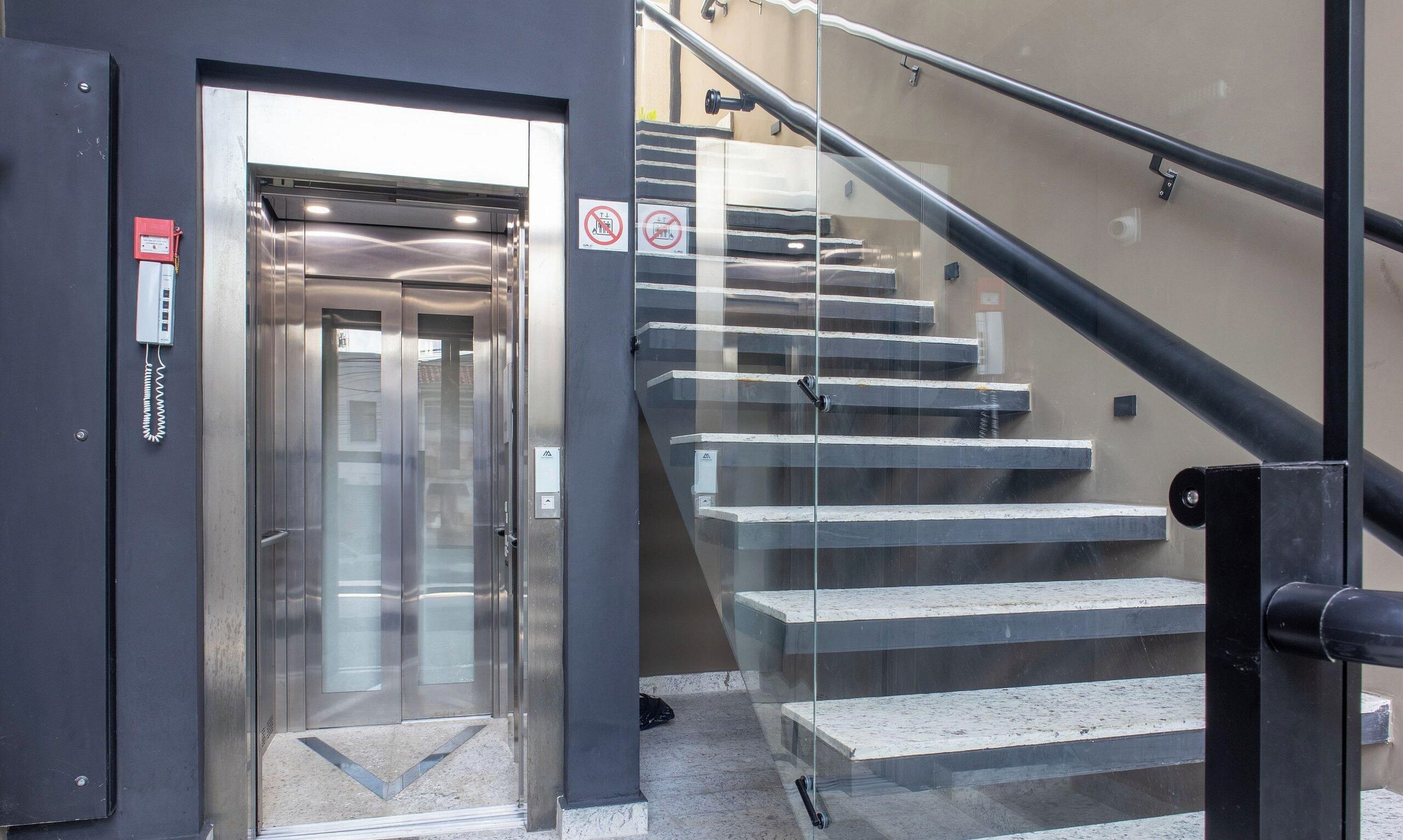Um elevador moderno com porta de vidro, um acesso por escadas com grades de metal e um painel de alarme de incêndio.