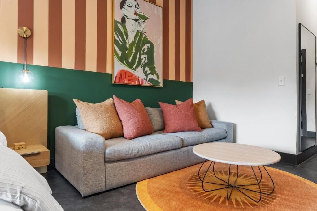 Quarto de hotel moderno com sofá cinzento, uma mesa de centro redonda e um quadro na parede.
