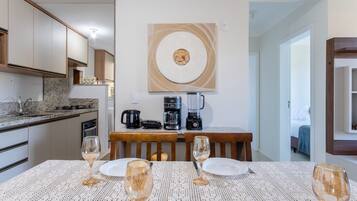 Uma área de jantar com uma mesa posta para quatro pessoas, uma cozinha ao fundo e uma cama visível através de uma porta aberta.