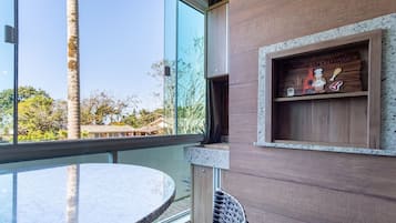 Quarto moderno com mesa de mármore redonda, uma cadeira de vime e uma prateleira de madeira com objetos decorativos. Uma janela grande oferece vista para árvores e um edifício lá fora.