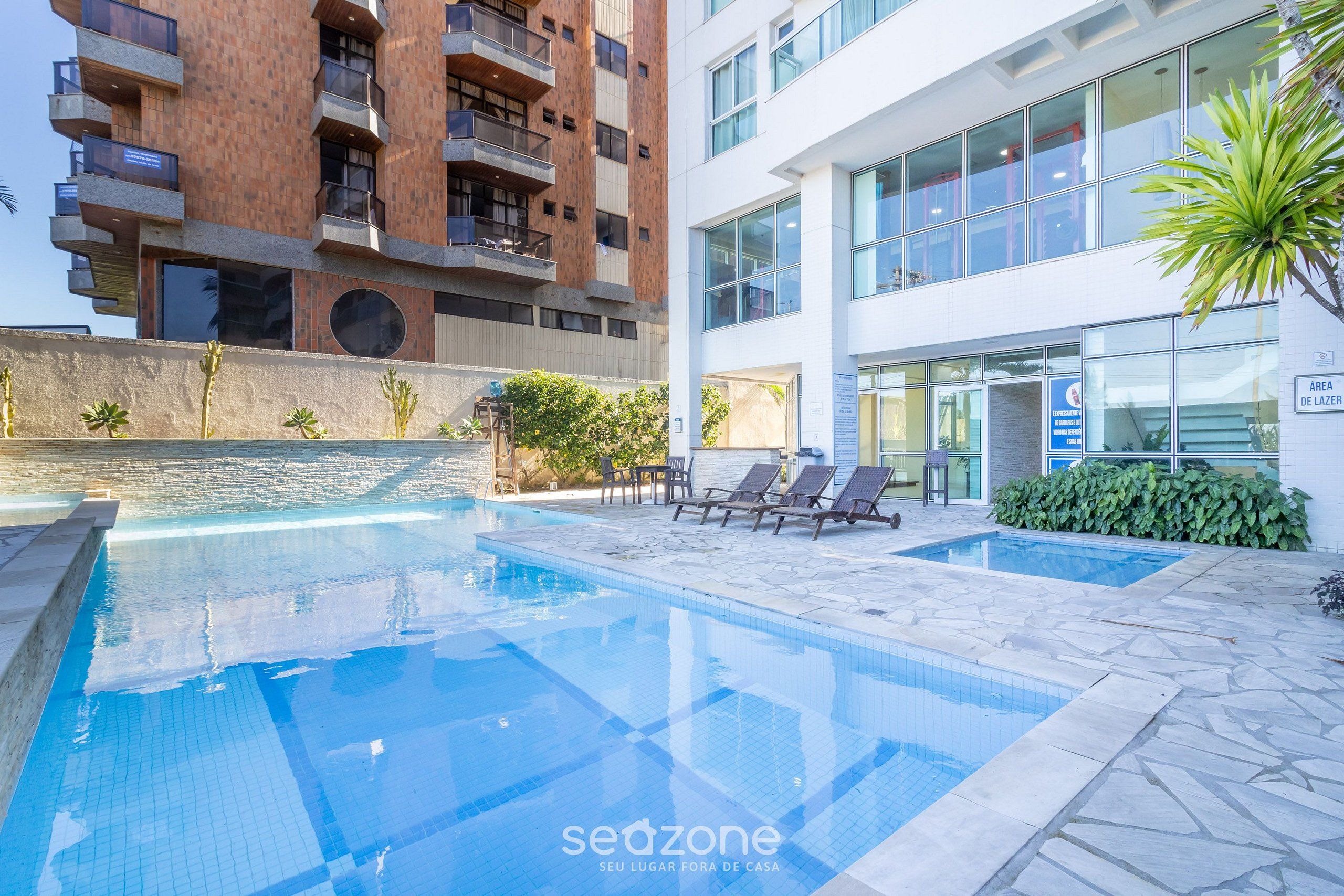 Uma área de piscina com espreguiçadeiras, um edifício com varandas e uma placa indicando espaço para atividades de lazer.