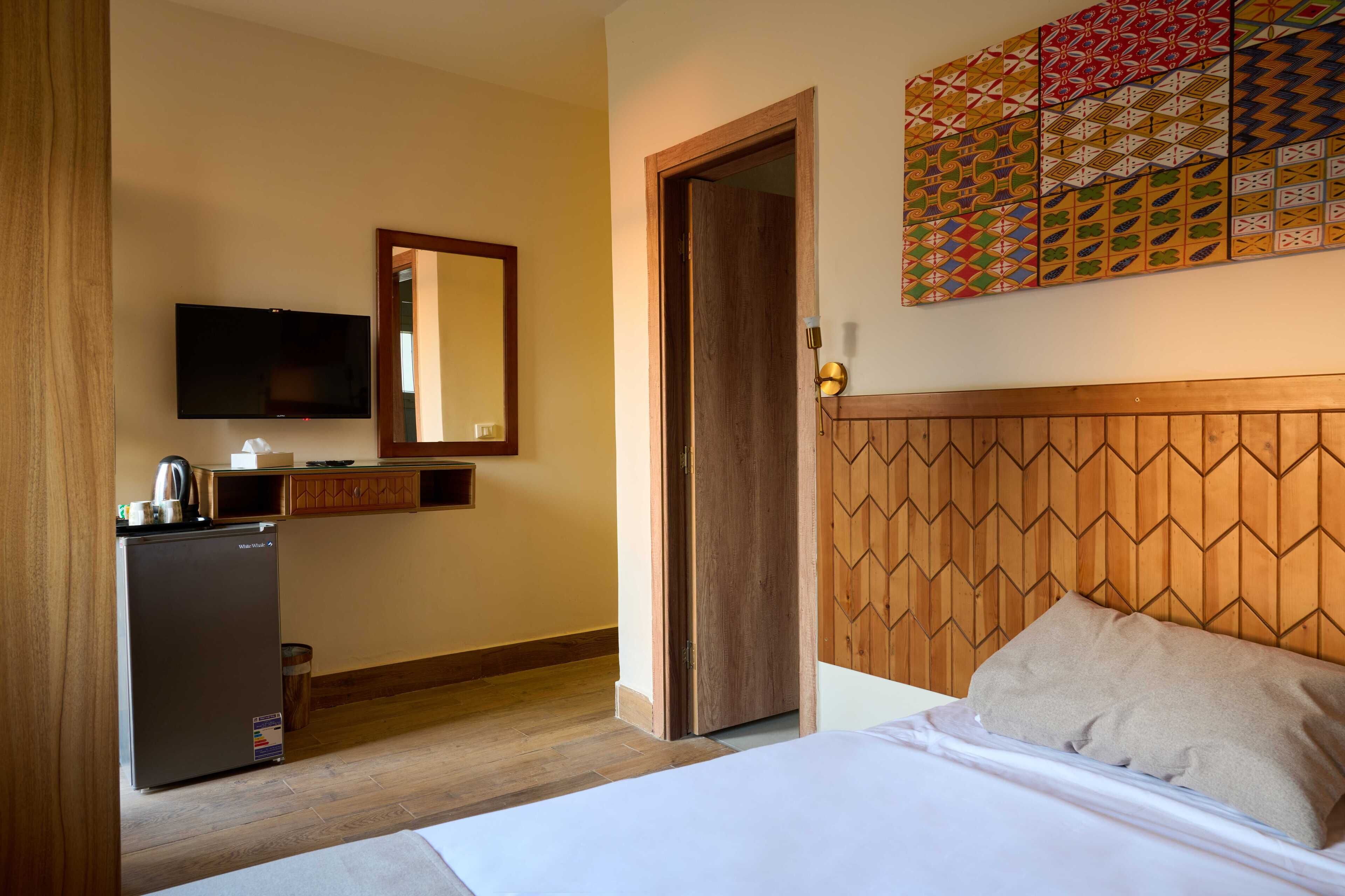 Quarto de hotel com cama, televisão, mini-frigorífico, cabeceira de madeira e parede com arte decorativa de padrão geométrico.